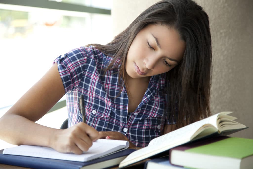 Student concentrating on writing in book with other books - Career Girls